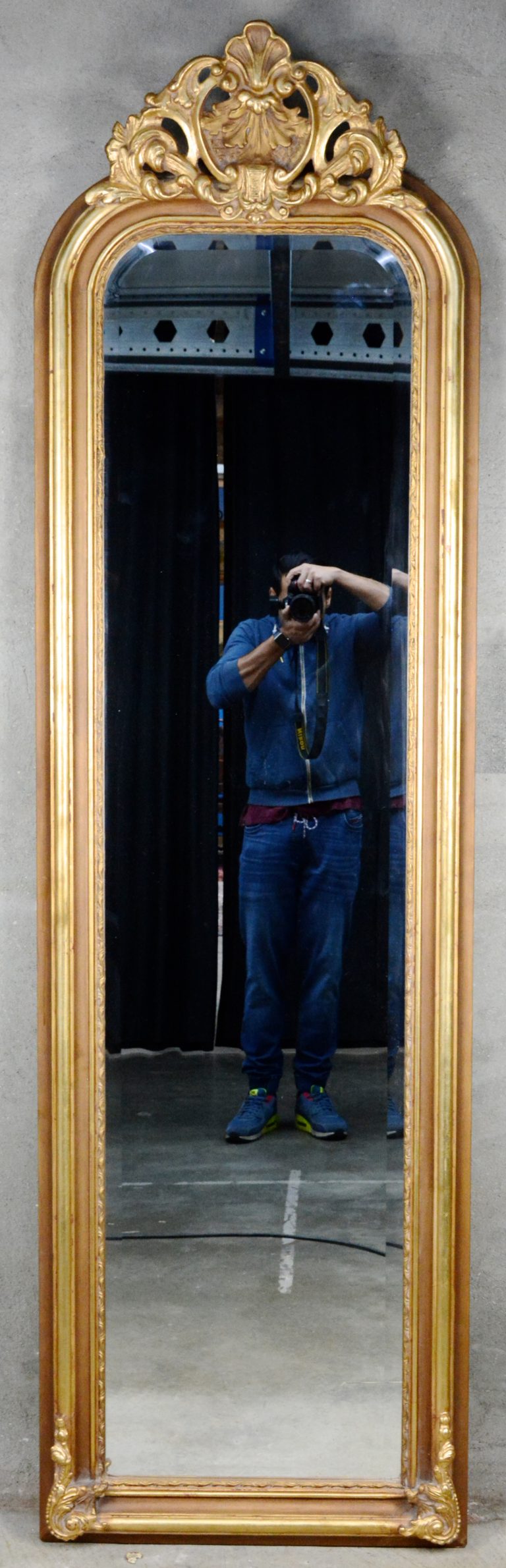 Een lange wandspiegel met verguld houten lijst, bovenaan versierd met ...